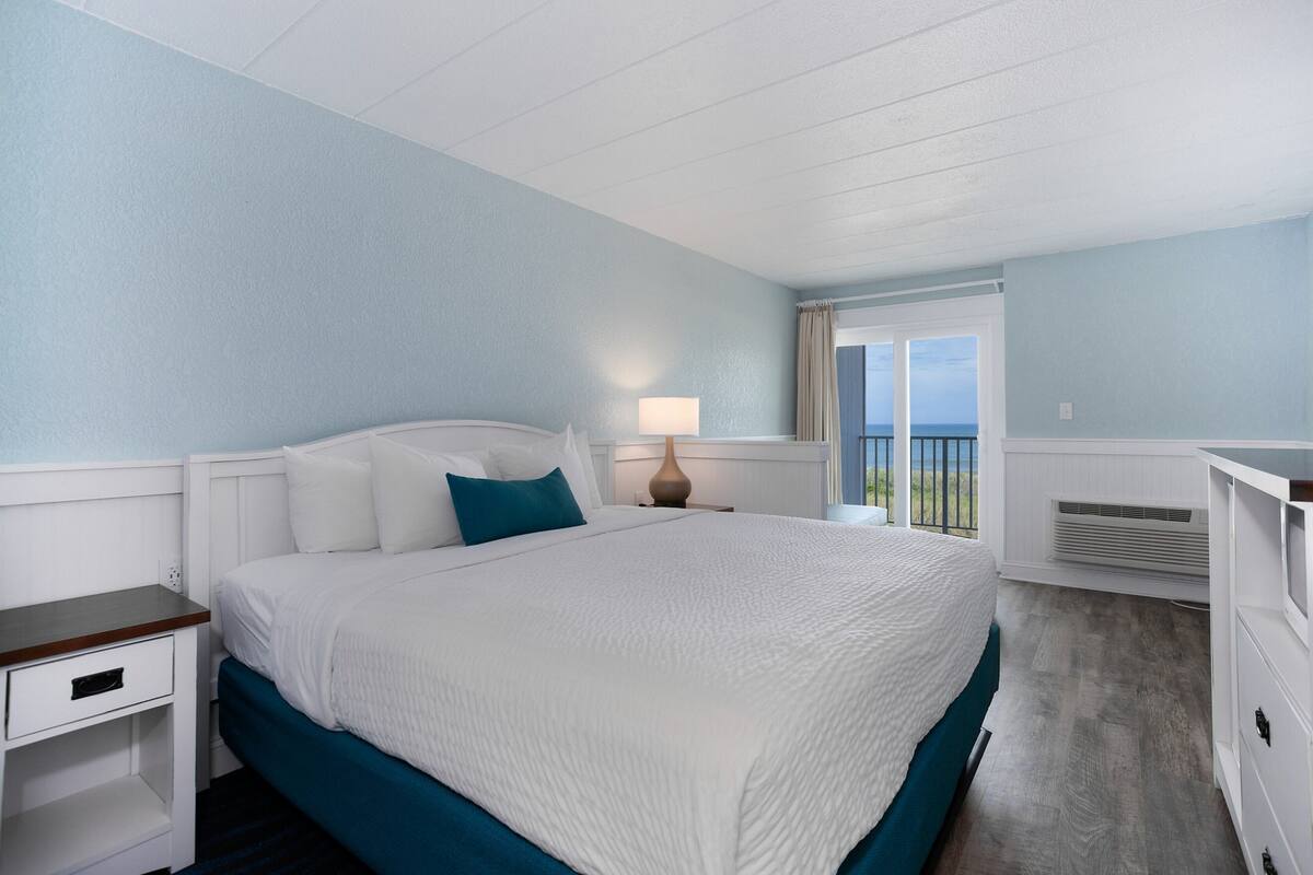 Quarto de hotel com uma cama grande, uma mesa de cabeceira, cômoda e vista para o oceano pela janela.