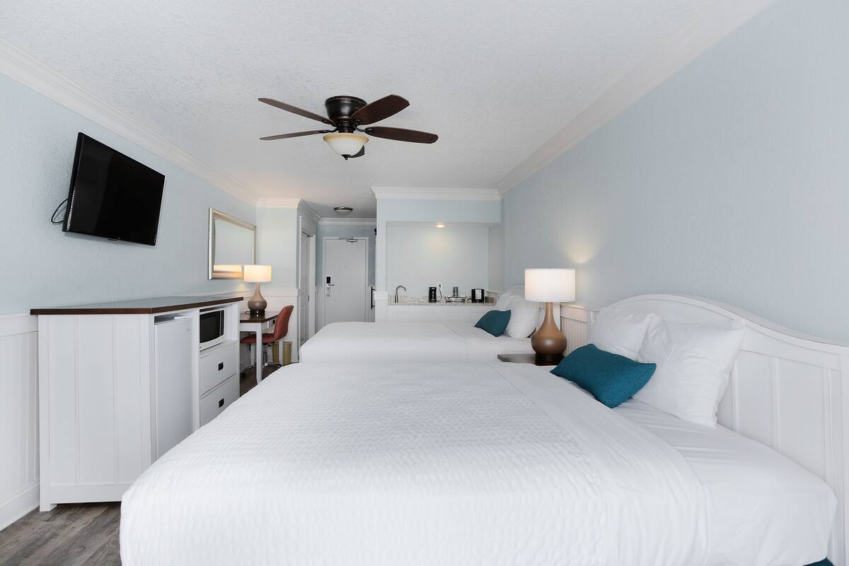 Quarto de hotel com uma cama grande, uma mesa de trabalho com uma TV, um ventilador de teto e um banheiro visível ao fundo.