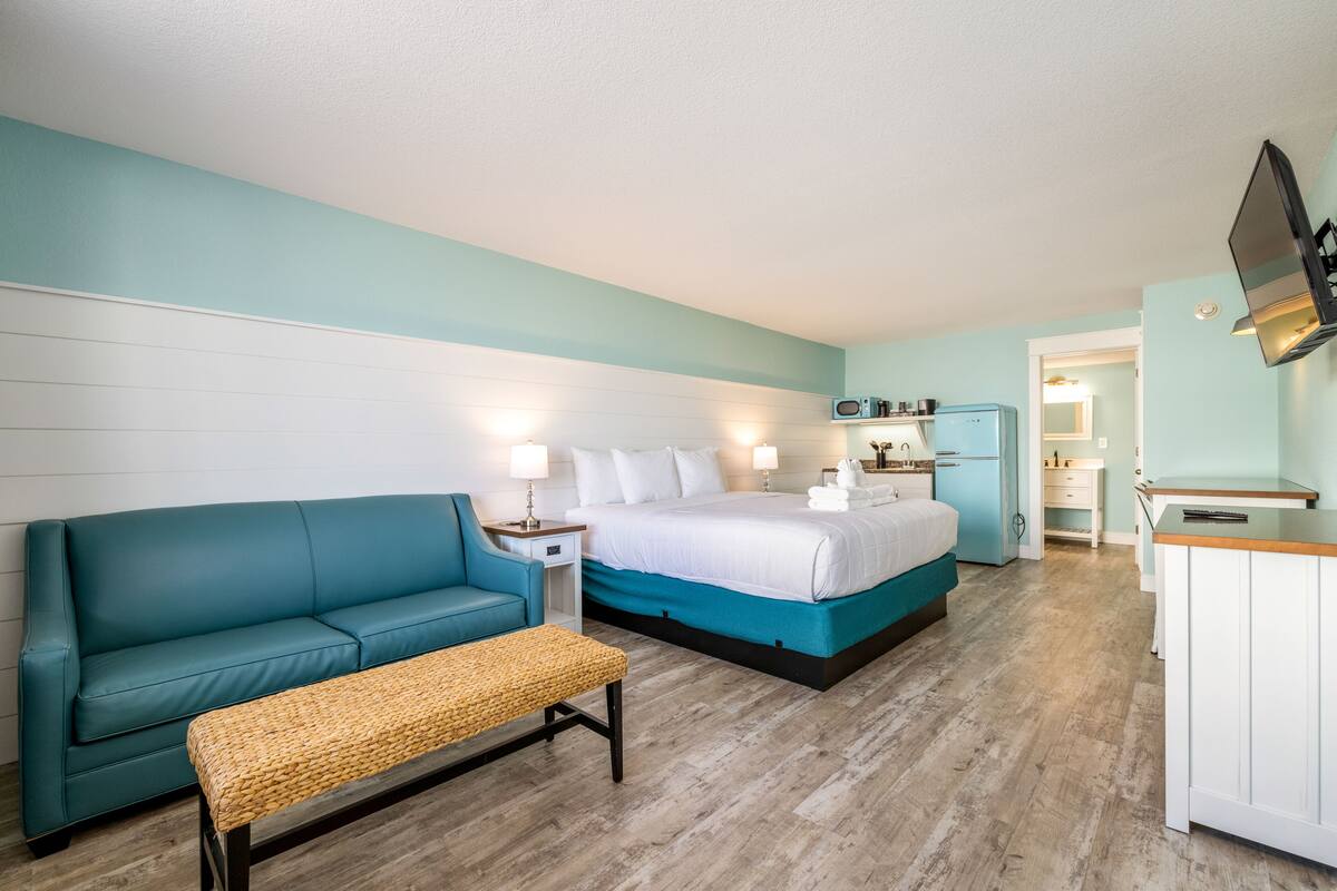 Quarto de hotel moderno com sofá verde-água, um banco de madeira, cama com roupa de cama branca, pequena cozinha e TV de tela plana na parede.