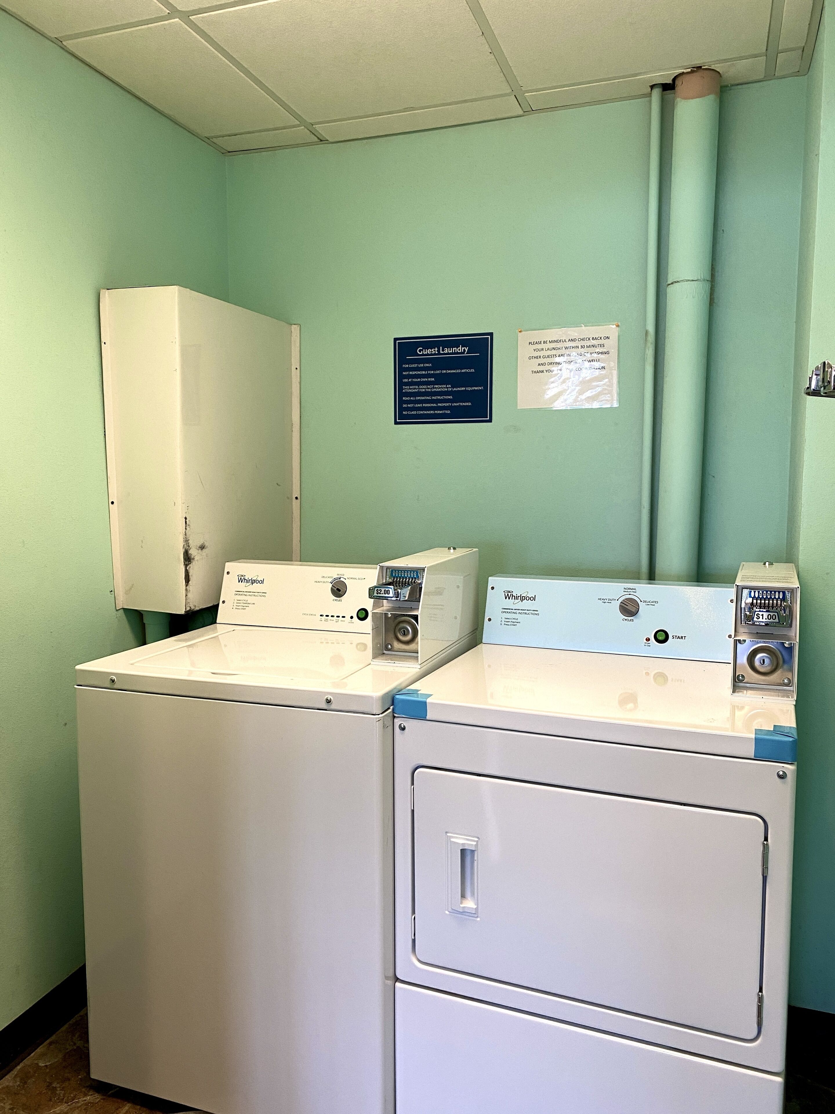 Uma lavanderia com duas máquinas de lavar e secar Whirlpool, uma placa indicando lavanderia para hóspedes e um aviso para verificar os itens de roupa.