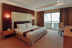 Quarto de hotel com uma cama grande, uma escrivaninha, uma cadeira, um vaso de plantas e vista para o exterior.