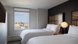 Quarto de hotel com duas camas, uma janela grande com vista para a cidade, uma luminária moderna e uma parede com padrão geométrico.