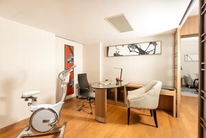 Quarto com bicicleta ergométrica, escrivaninha, cadeira e obras de arte nas paredes.