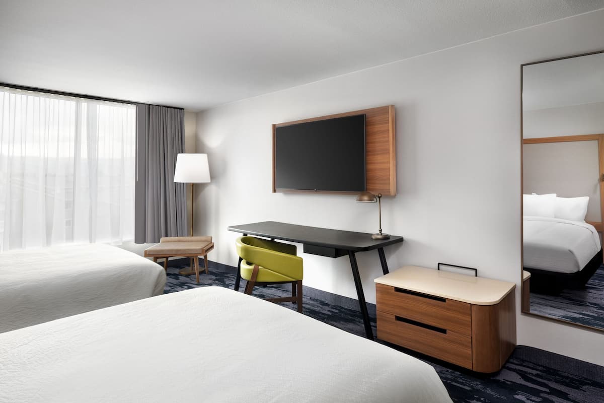 Quarto de hotel com duas camas, uma escrivaninha, uma cadeira, uma televisão e um espelho.