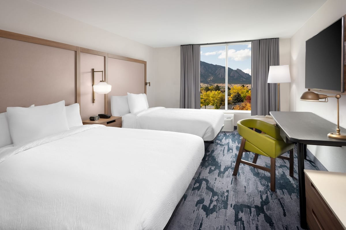 Quarto de hotel com duas camas, uma escrivaninha e uma cadeira. Há uma janela grande com vista para as montanhas.