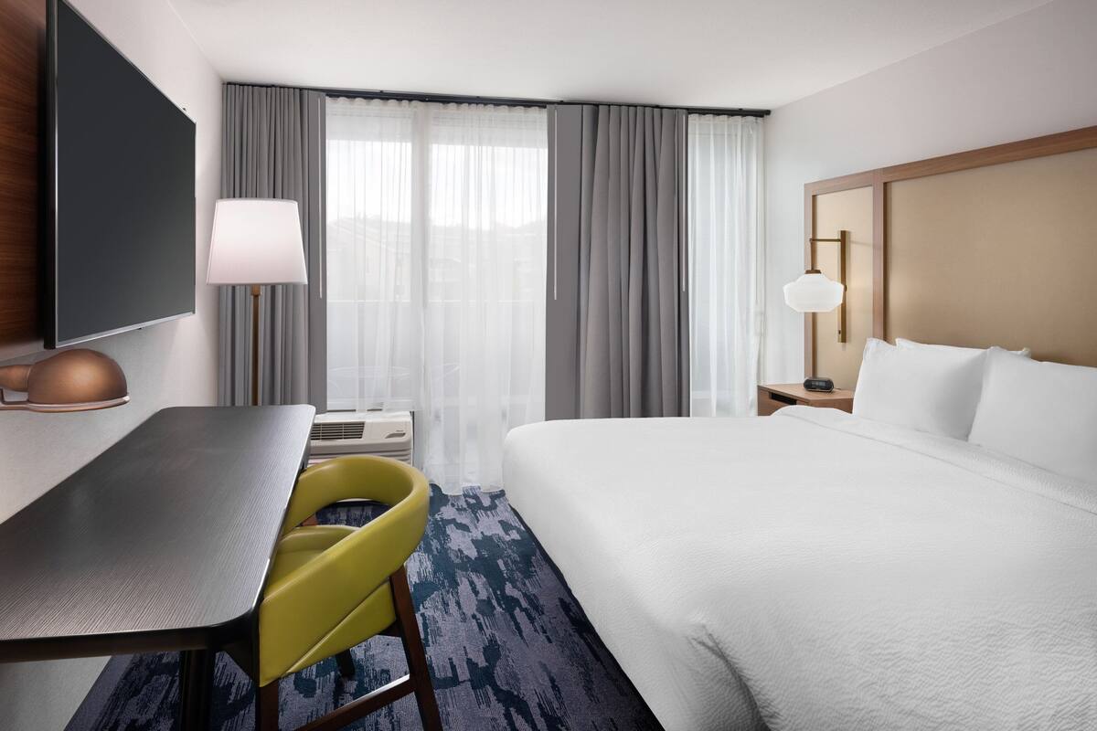 Quarto de hotel com cama, escrivaninha, cadeira, televisão, luminária e janela com cortinas.