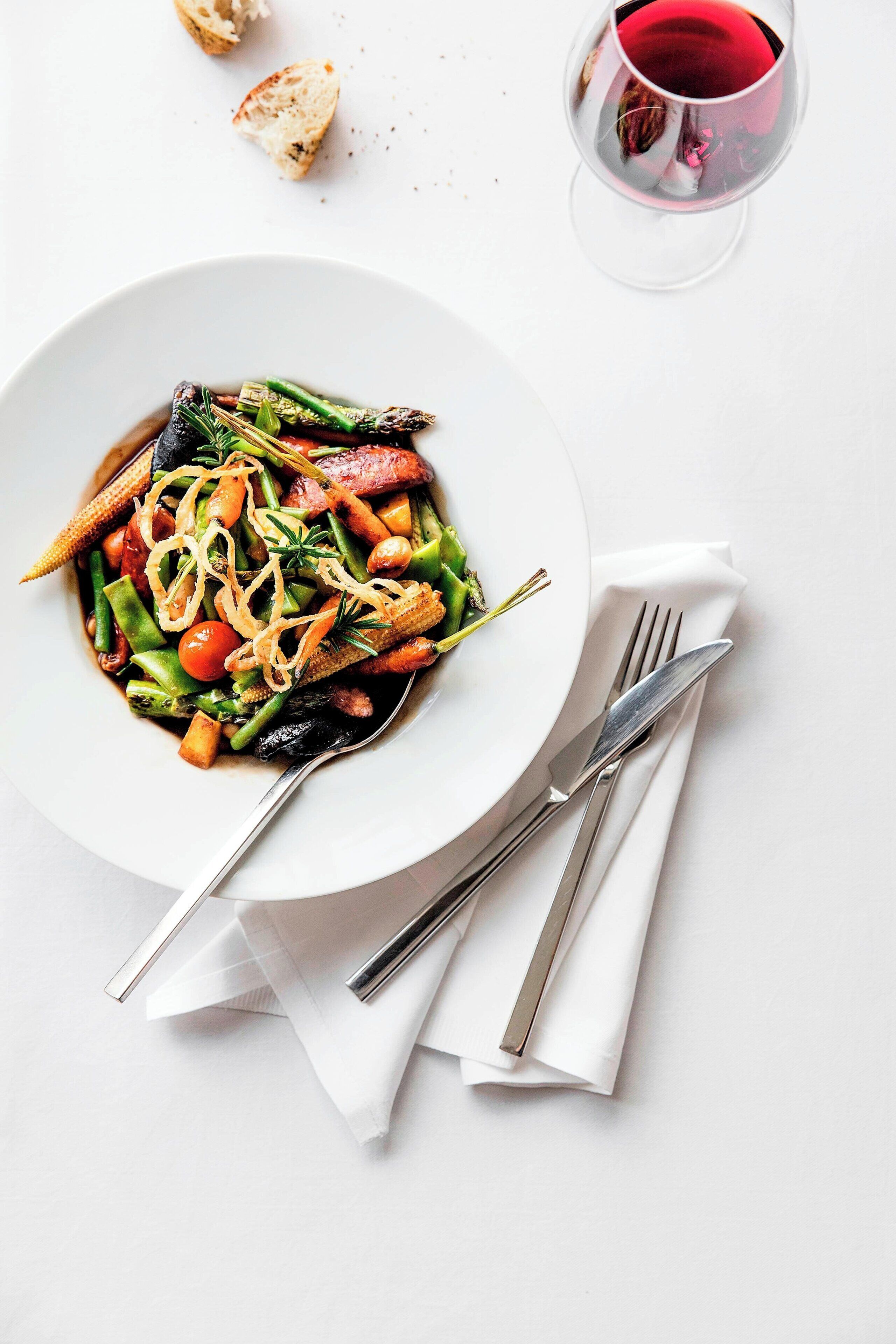 Um prato branco com um prato de legumes, um garfo, uma faca e um copo de vinho tinto sobre uma mesa branca.