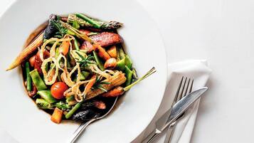 Um prato branco com um prato de legumes, um garfo, uma faca e um copo de vinho tinto sobre uma mesa branca.