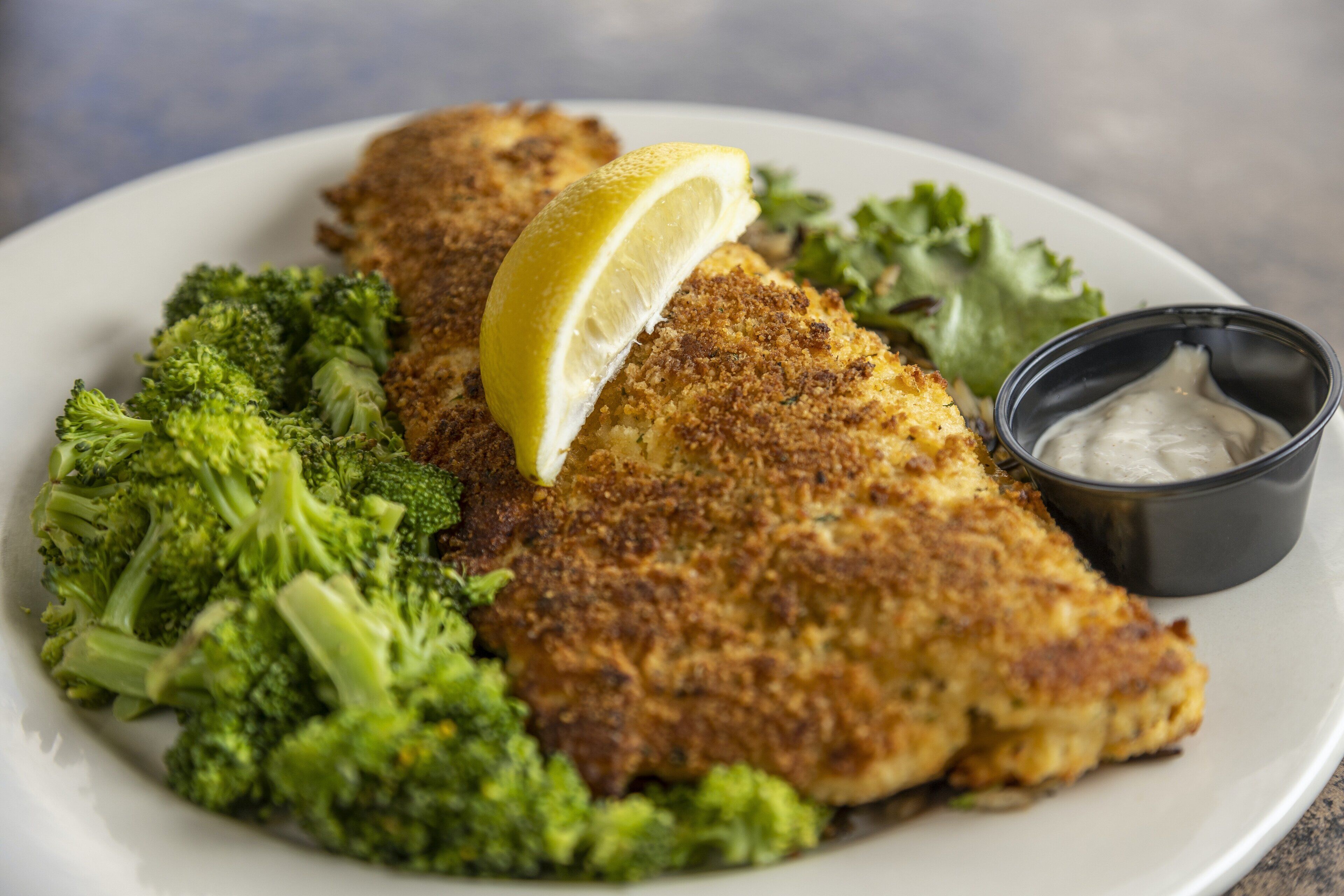 Um prato com escalope à milanesa, um pedaço de limão, brócolis e uma pequena tigela de molho.