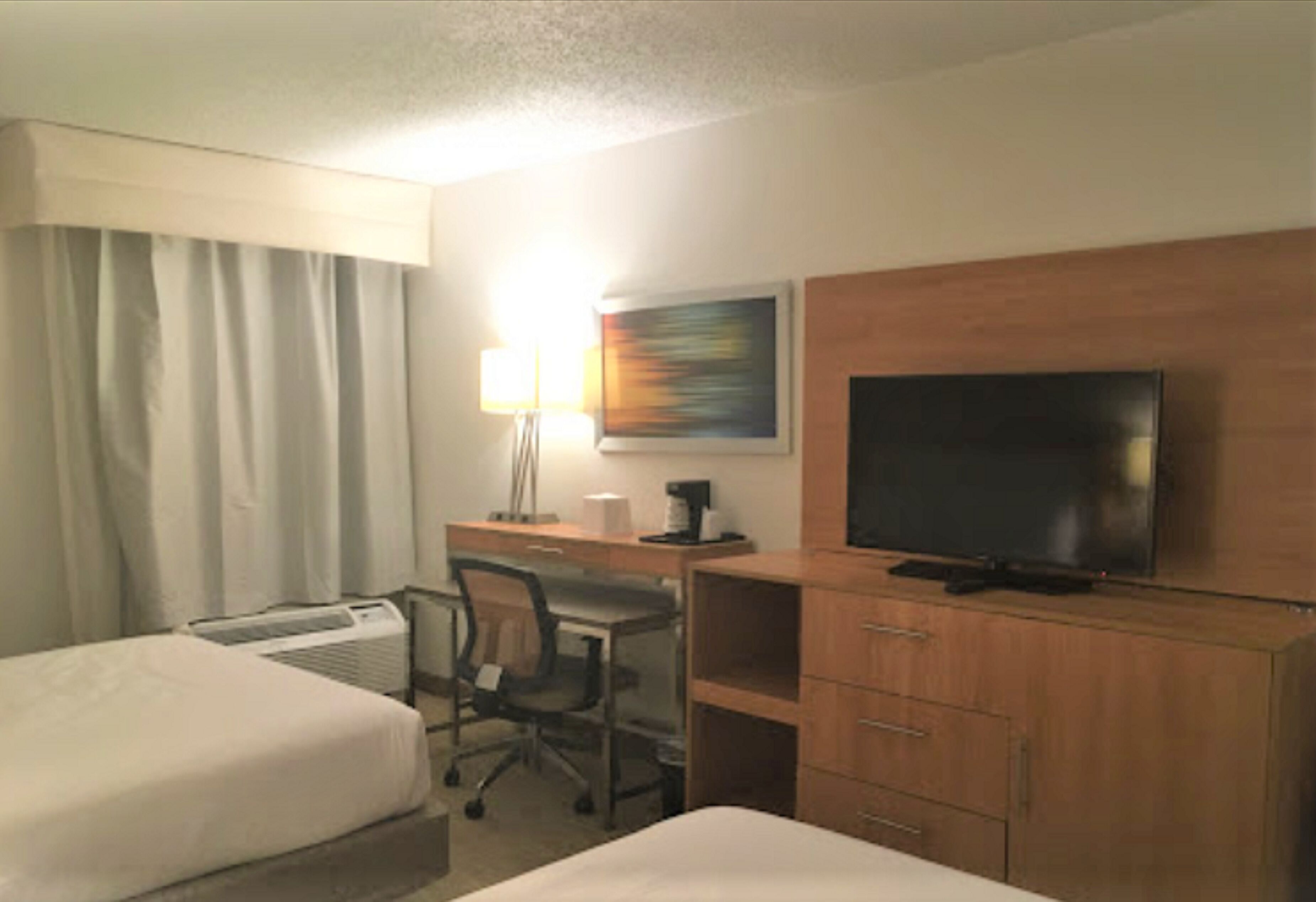 Quarto de hotel com duas camas, uma escrivaninha, uma cadeira, uma televisão e um quadro na parede.