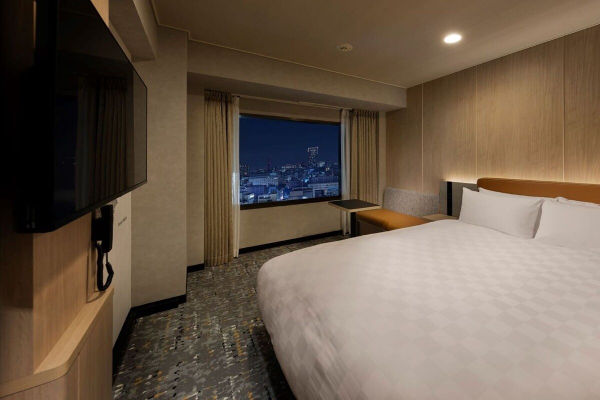 Quarto de hotel com cama, televisão, escrivaninha, cadeira, janela com vista para a cidade e parede de madeira.