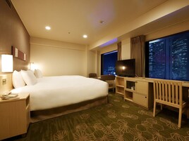 Quarto de hotel com uma cama grande, uma escrivaninha com televisão, vista para a cidade e um tapete com padrão geométrico.