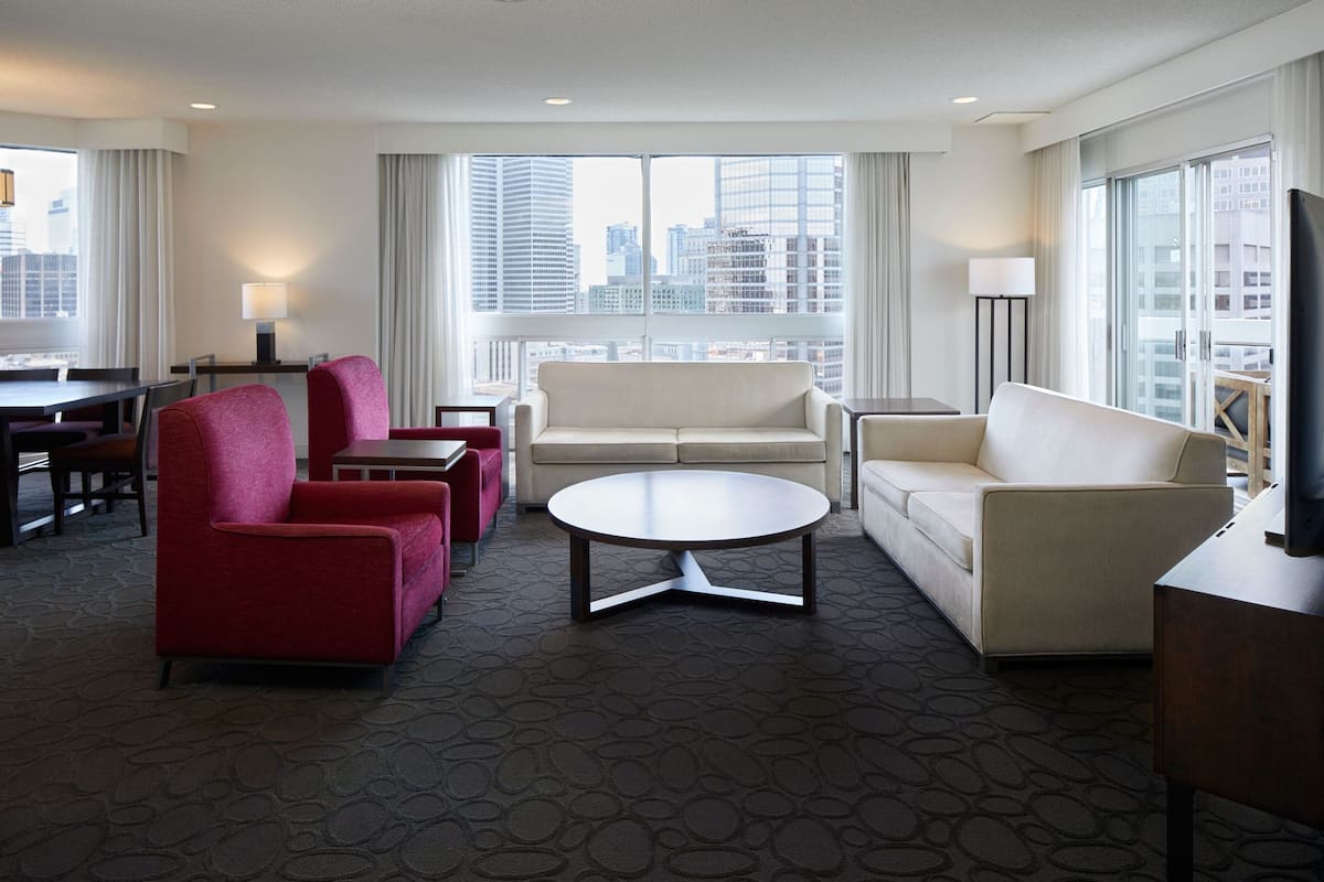 Quarto de hotel com uma janela grande, um sofá, uma poltrona vermelha, uma mesa de centro redonda e uma TV sobre um suporte.