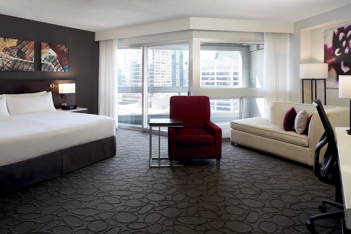 Quarto de hotel com uma cama grande, uma poltrona vermelha, um sofá, uma escrivaninha e uma janela ampla com vista para a cidade.