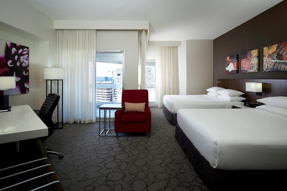 Quarto de hotel com duas camas, uma escrivaninha, uma cadeira e vista para a paisagem urbana pela janela.