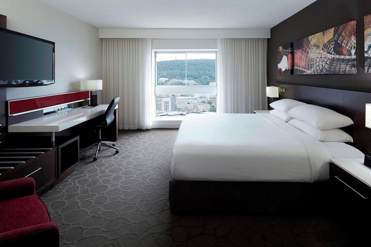 Quarto de hotel com uma cama grande, uma mesa com cadeira, uma televisão e uma janela com cortinas.