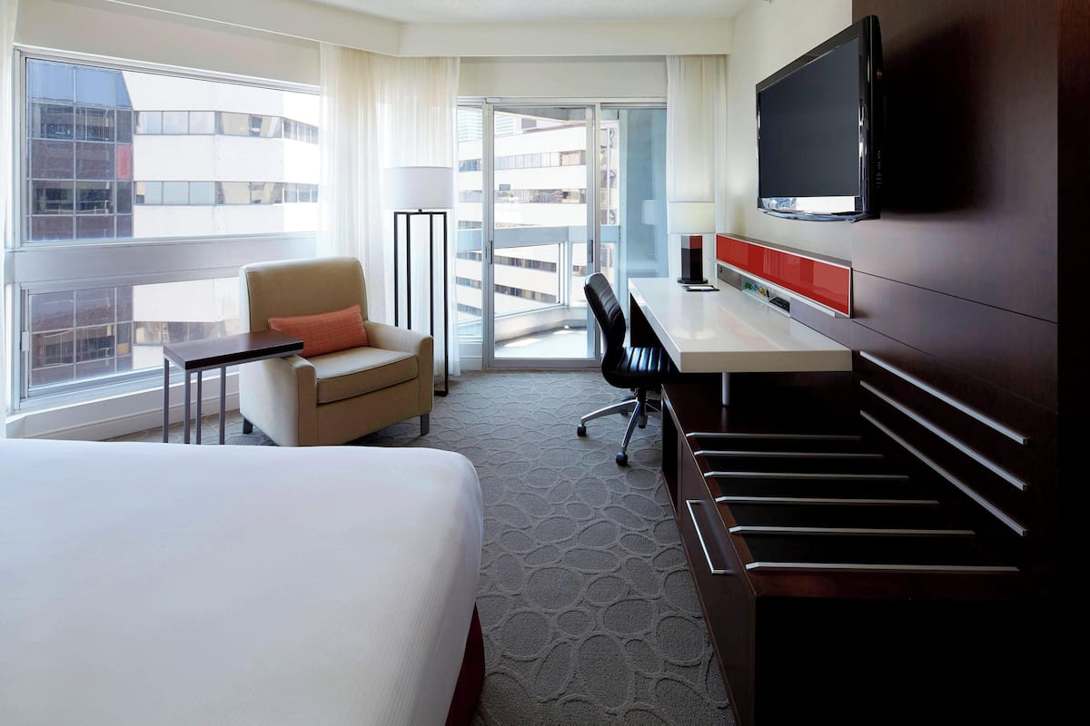 Quarto de hotel com cama, escrivaninha, cadeira, televisão e vista da cidade através de amplas janelas.