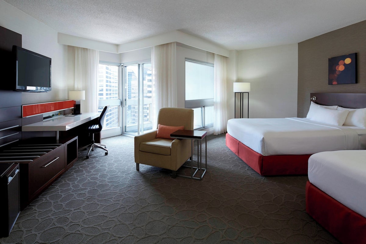 Quarto de hotel com cama, escrivaninha, cadeira, televisão e janela com cortinas.