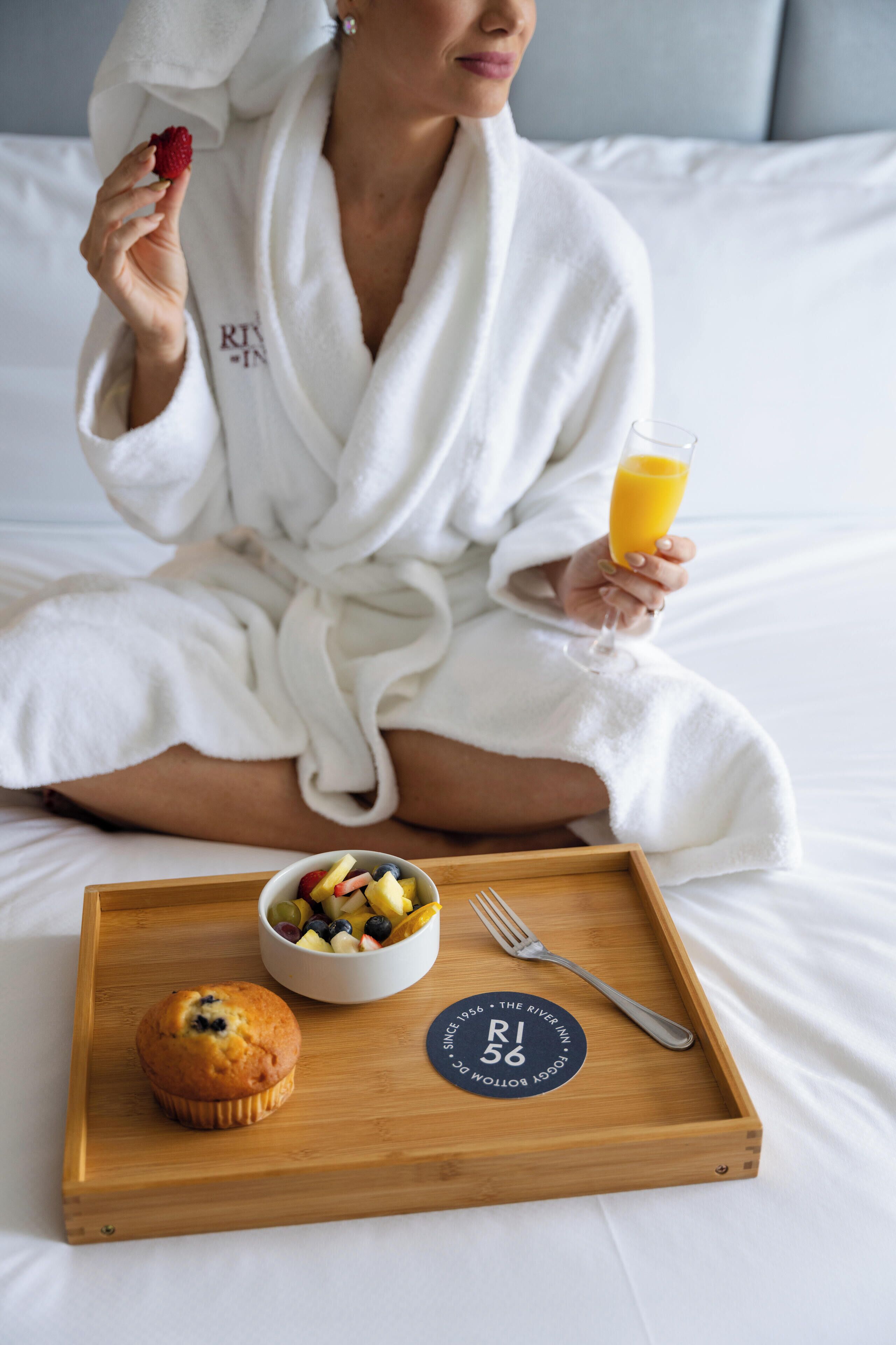 Uma pessoa vestindo um roupão branco sentada na cama com uma bandeja contendo um muffin, uma tigela de frutas e um copo de suco de laranja.