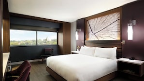 Quarto de hotel com uma cama grande, uma escrivaninha, uma cadeira, janela com vista e uma obra de arte na parede.