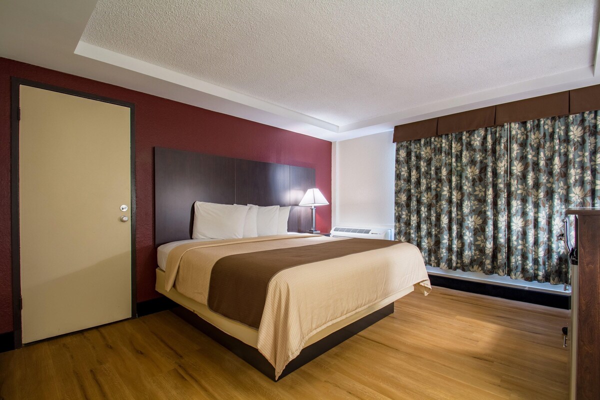 Quarto de hotel com cama, mesa de cabeceira, abajur, janela com cortinas e porta.