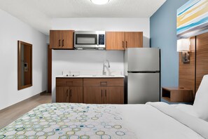 Quarto de hotel compacto com cozinha americana, uma cama com colcha estampada e uma mesa de cabeceira de madeira.