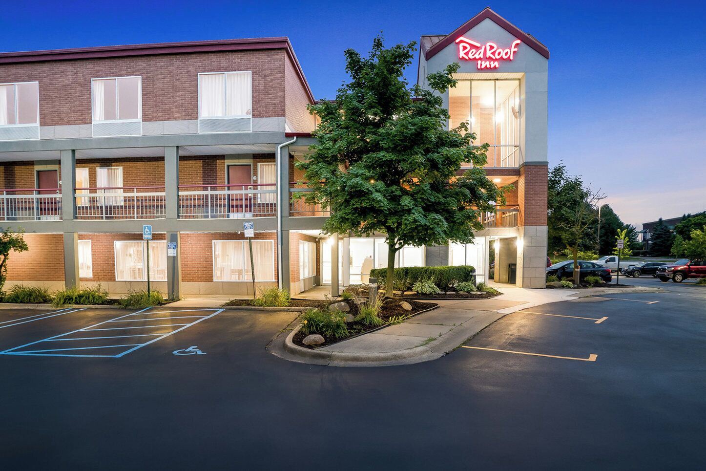 Um Red Roof Inn com um edifício de dois andares, estacionamento e área ajardinada com uma árvore.