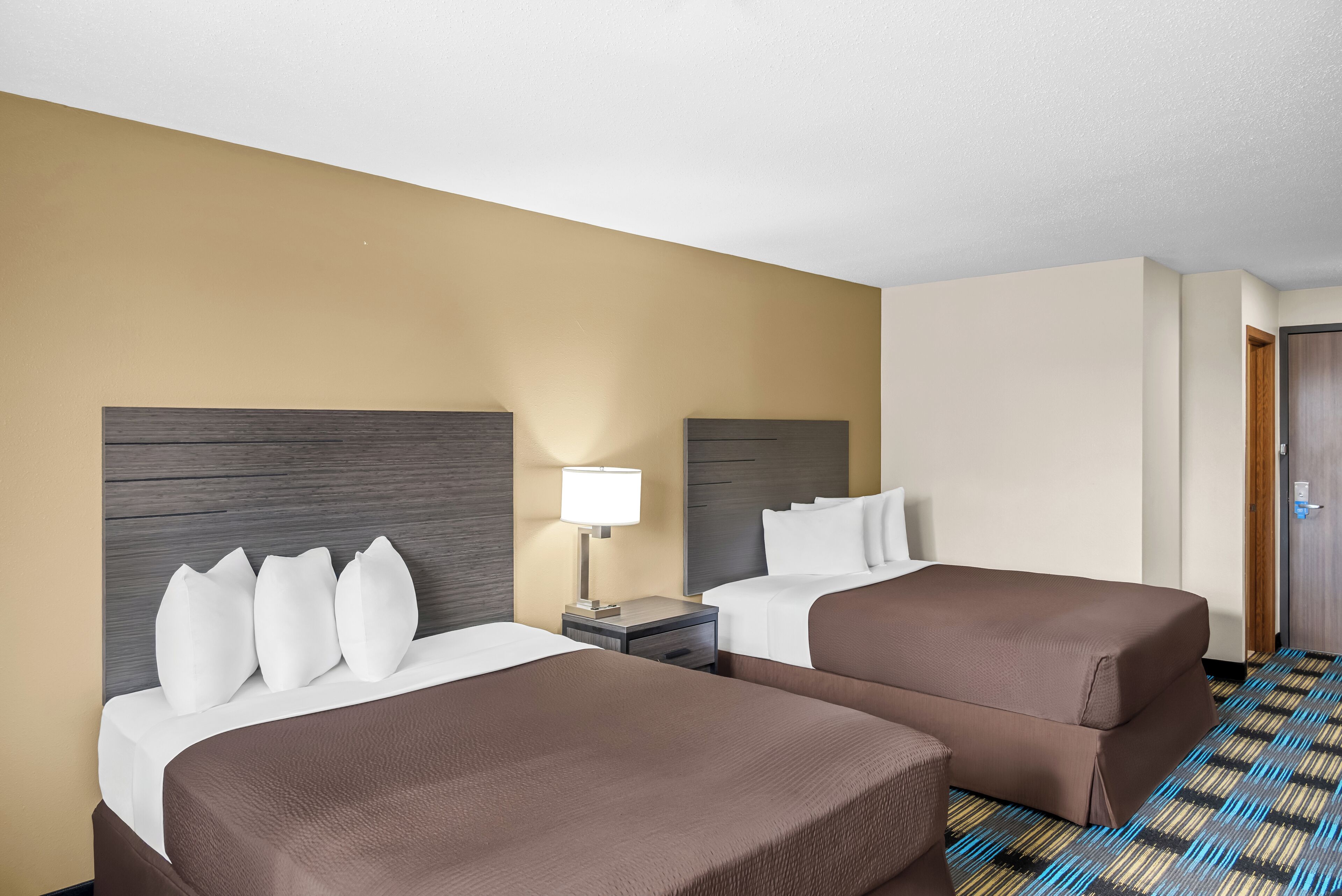Quarto de hotel com duas camas, uma mesinha de cabeceira com abajur e uma porta.