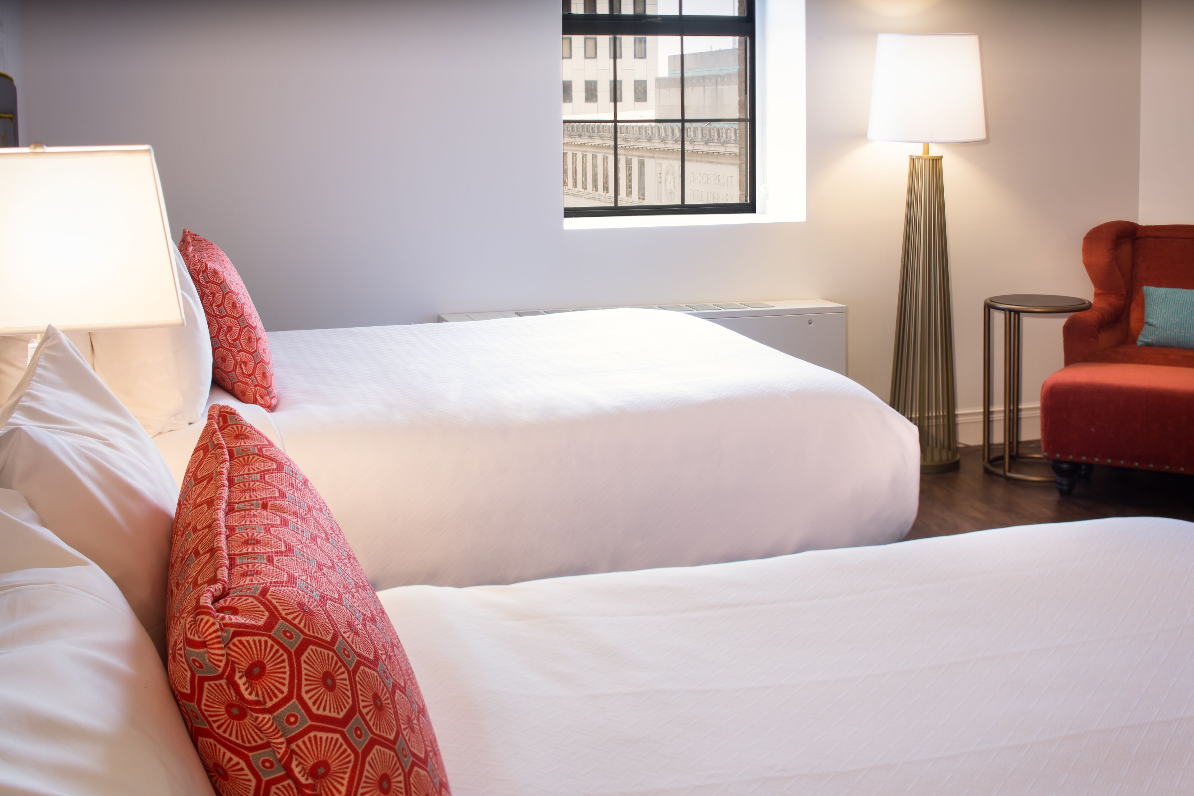 Quarto de hotel com duas camas, uma cadeira vermelha, abajur e janela com vista para a cidade.