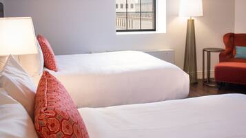 Quarto de hotel com duas camas, uma cadeira vermelha, abajur e janela com vista para a cidade.