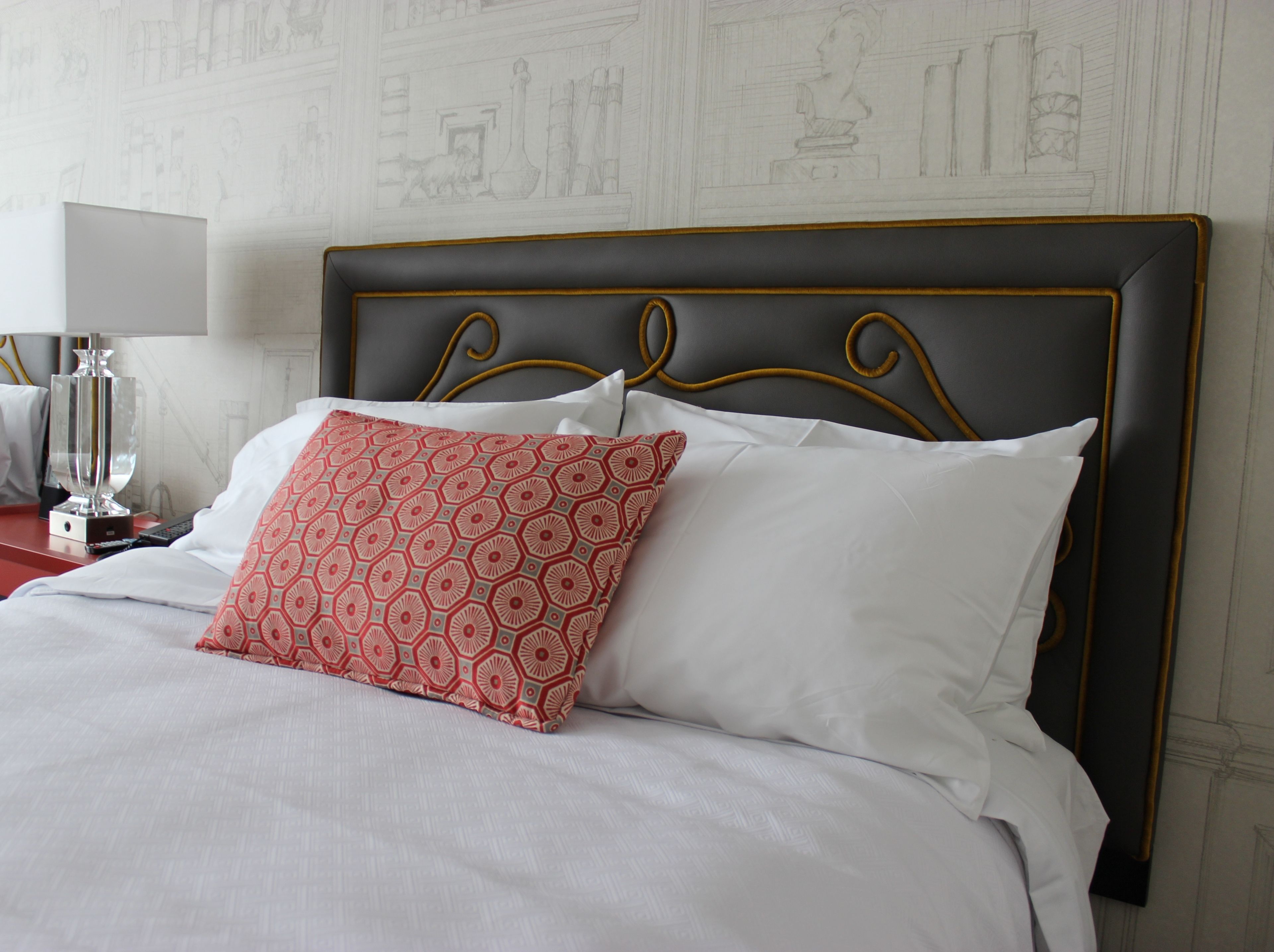 Uma cama bem arrumada com almofadas estampadas em branco e vermelho, uma cabeceira escura e uma mesa de cabeceira com luminária e controle remoto.