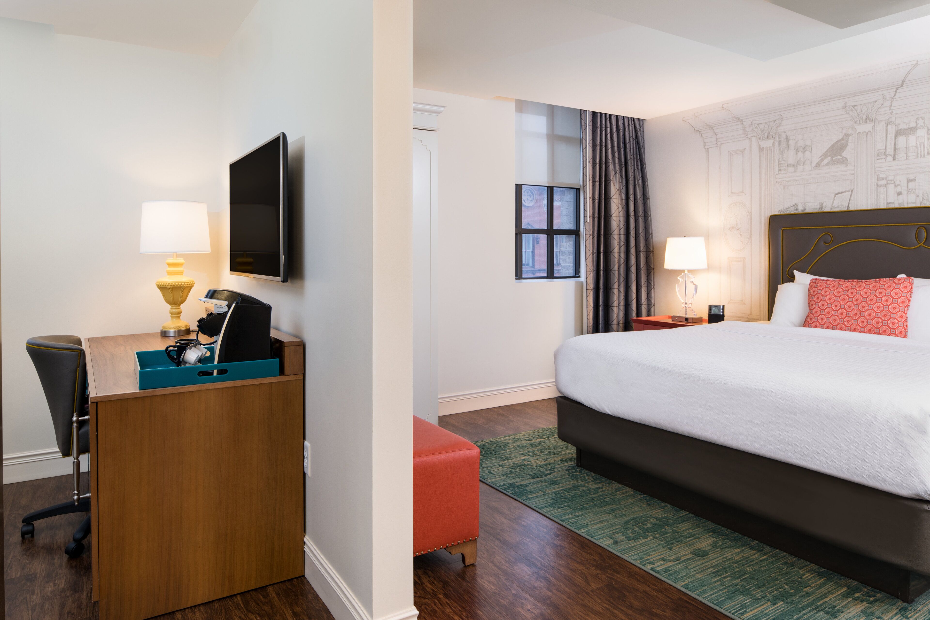 Quarto de hotel com cama, escrivaninha com cadeira, televisão e janela com cortinas.