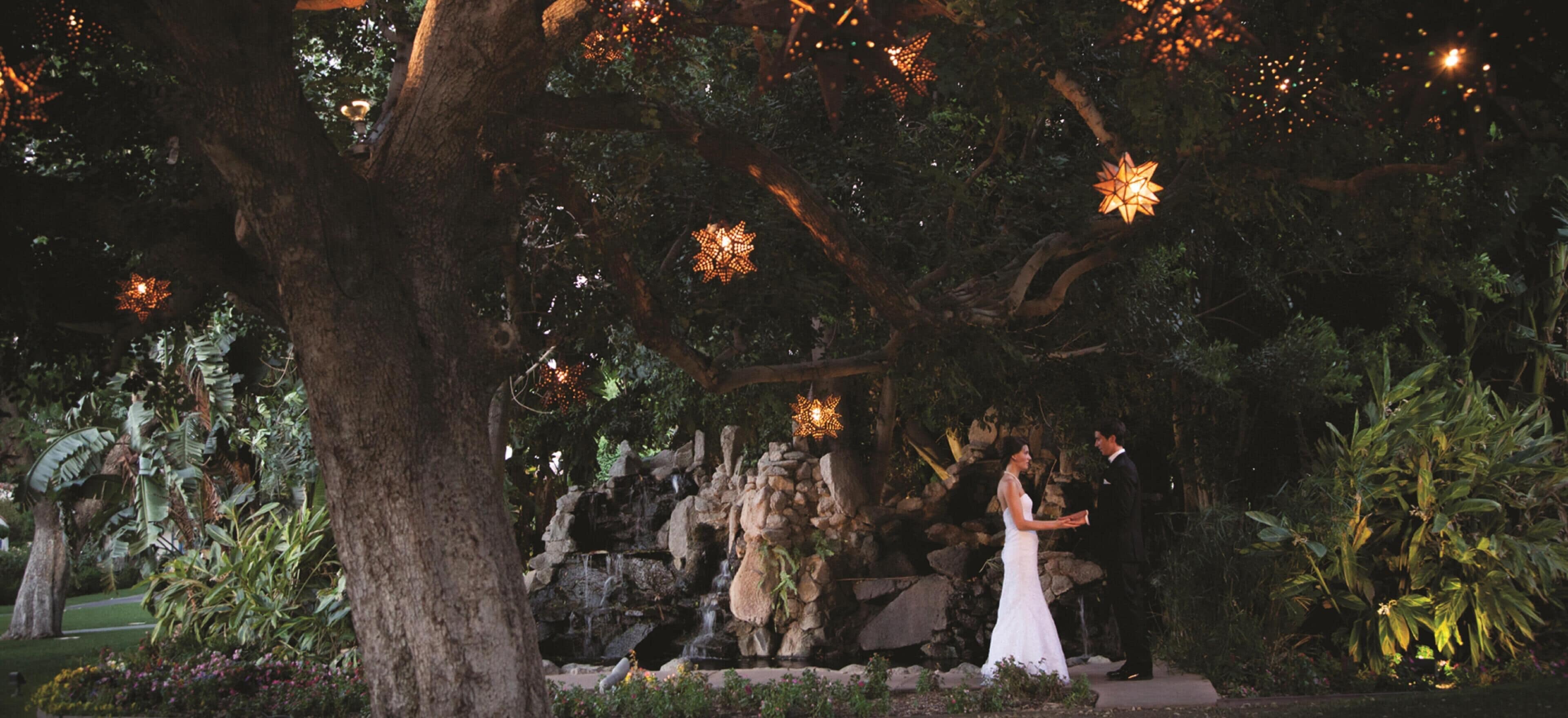 Um casal de mãos dadas em um jardim com uma cachoeira e luzes decorativas.