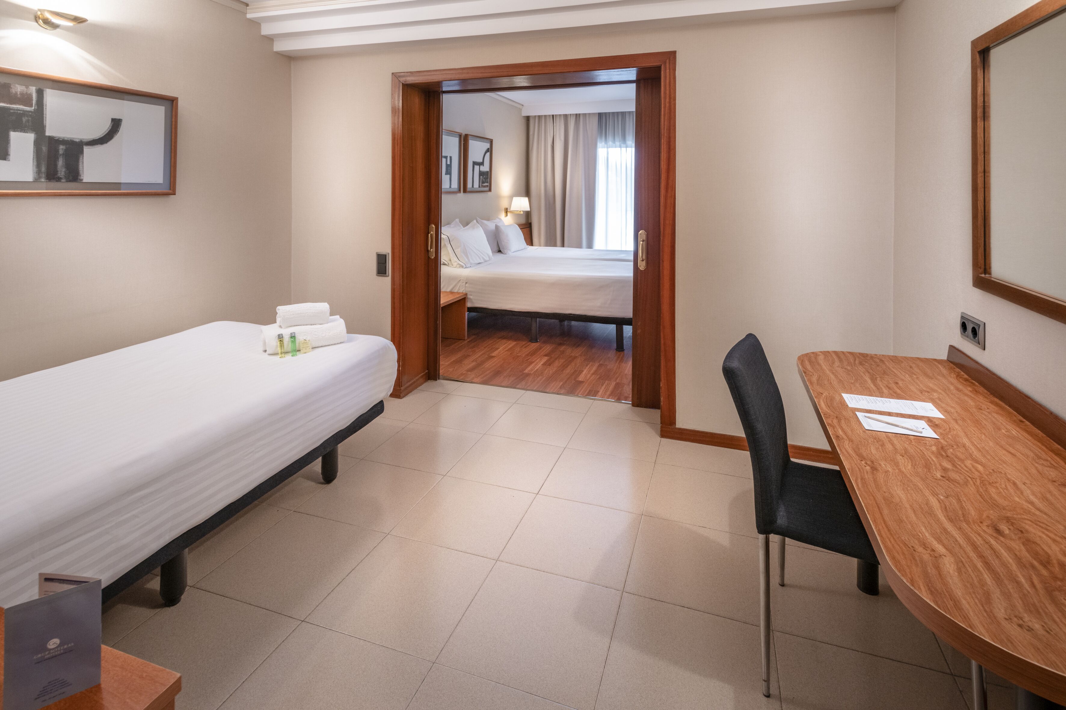 Quarto de hotel com uma cama de solteiro, uma escrivaninha de madeira, uma cadeira e uma pequena mesa com abajur.