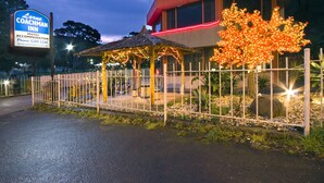 Um motel com uma placa, uma área cercada com uma árvore decorada com luzes, e um edifício com fachada iluminada em vermelho.