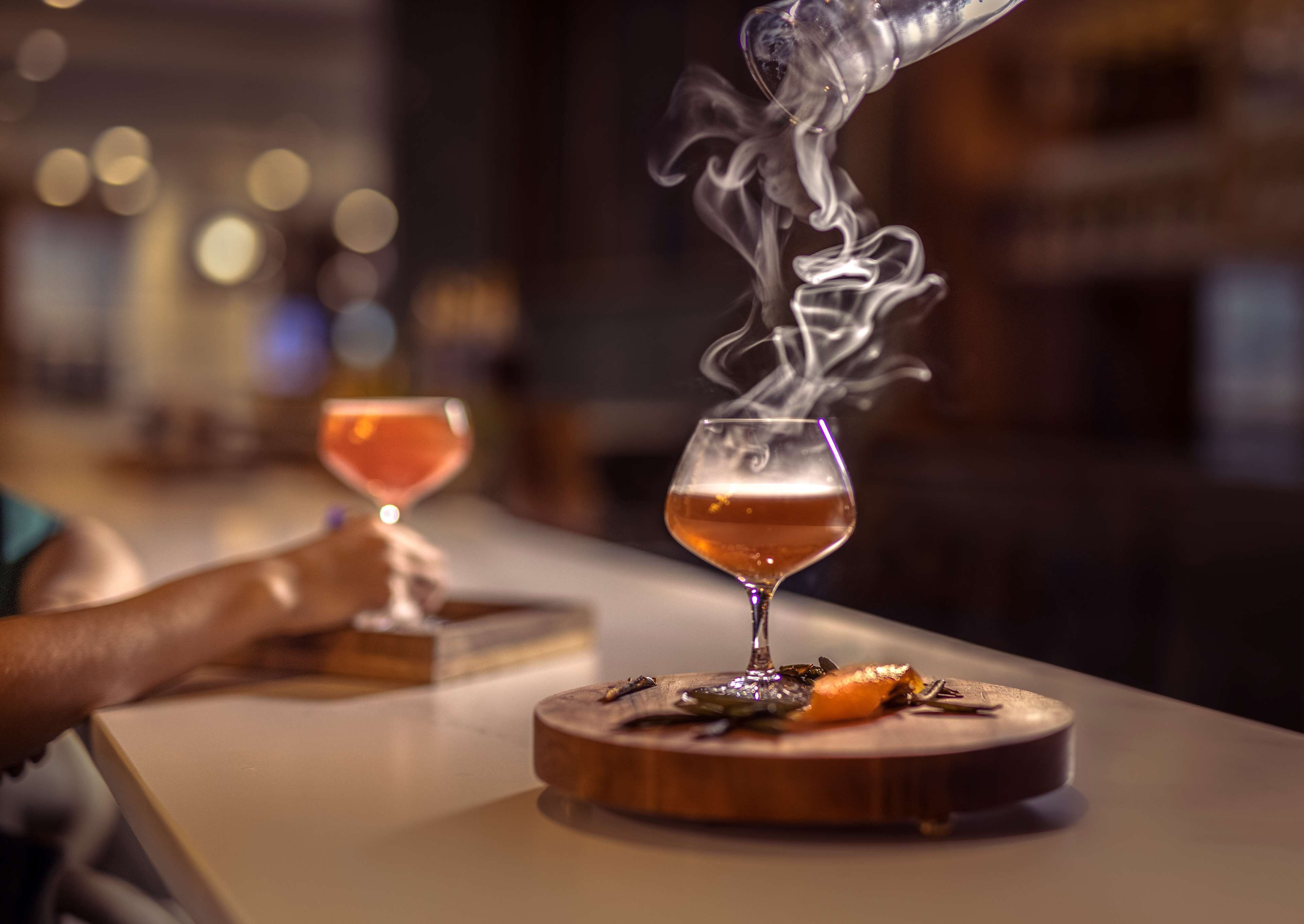 Um balcão de bar com um copo de bebida âmbar e um copo esfumado sobre um apoio de madeira.