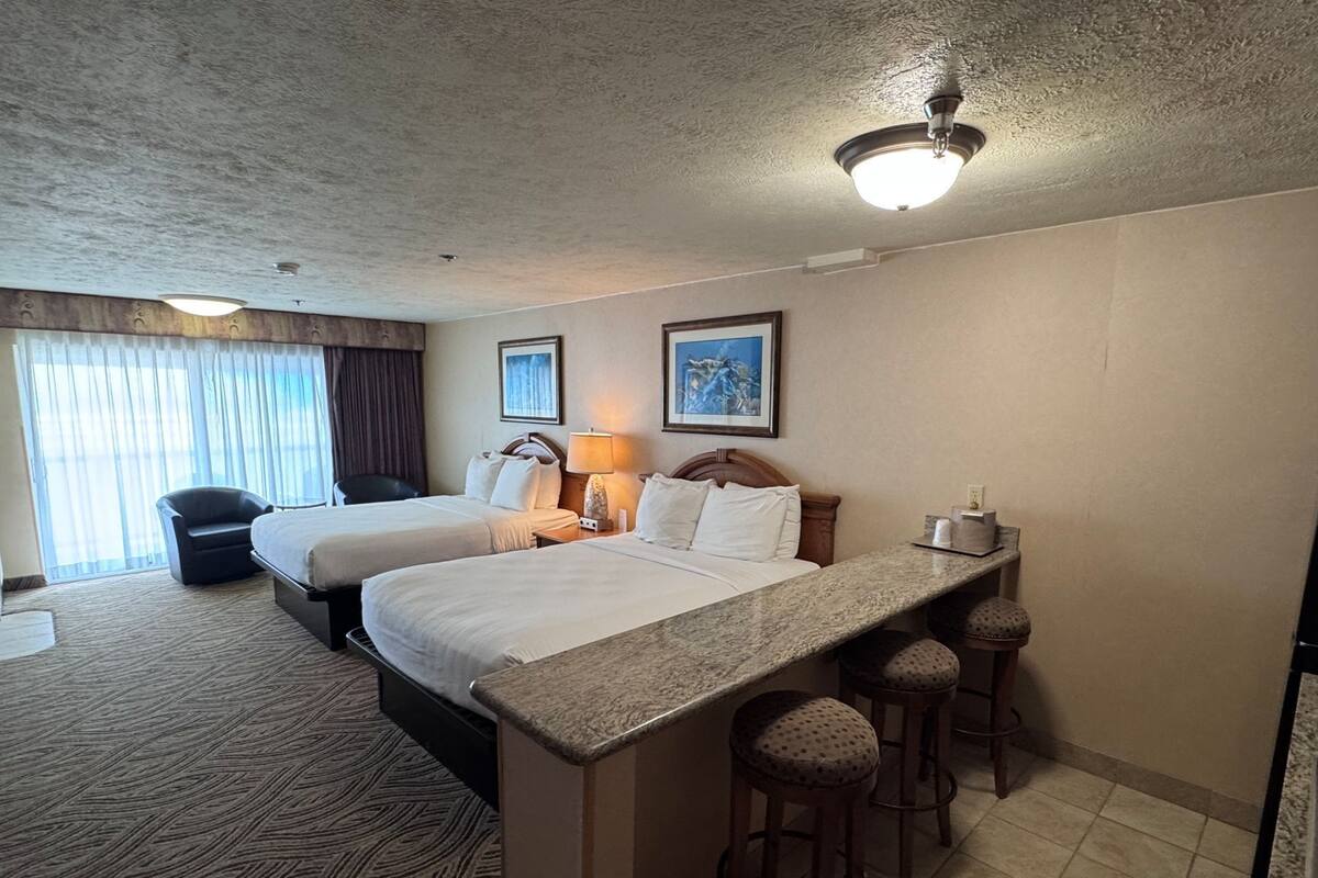 Quarto de hotel com duas camas, uma mesa com bancos, uma cadeira e uma janela grande com cortinas.