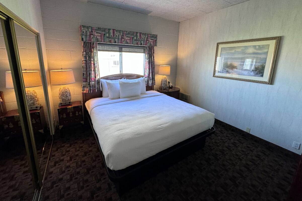 Quarto de hotel com uma cama grande, duas luminárias de cabeceira, mesa de cabeceira, espelho, quadro pendurado na parede e janela com cortinas.