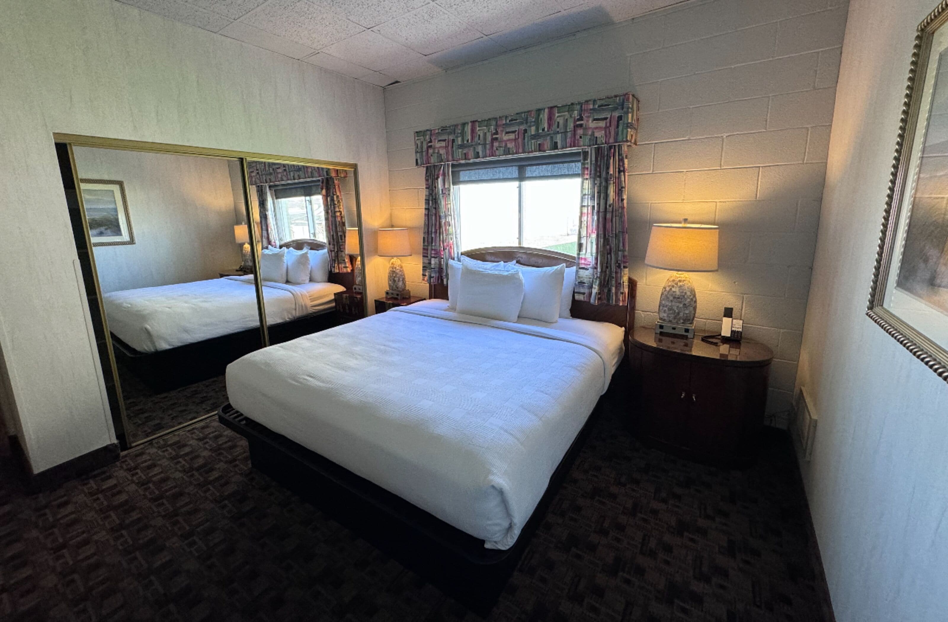 Quarto de hotel com uma cama grande, um espelho, uma janela com cortinas e uma mesa de cabeceira com abajur.
