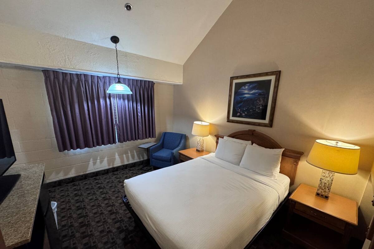 Quarto de hotel com uma cama grande, uma escrivaninha, uma cadeira, duas luminárias de cabeceira, uma janela com cortinas e um quadro na parede.