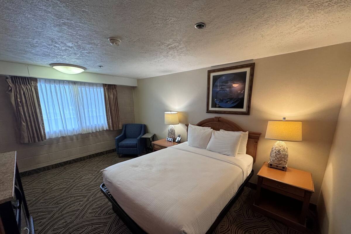 Quarto de hotel com uma cama grande, duas mesas de cabeceira com luminárias, uma cadeira, uma escrivaninha e uma janela com cortinas.