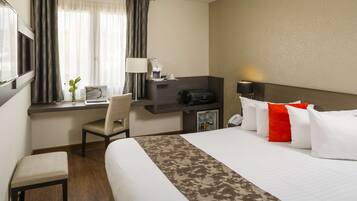 Quarto de hotel com cama, escrivaninha, cadeira, pequeno frigorífico e televisão.