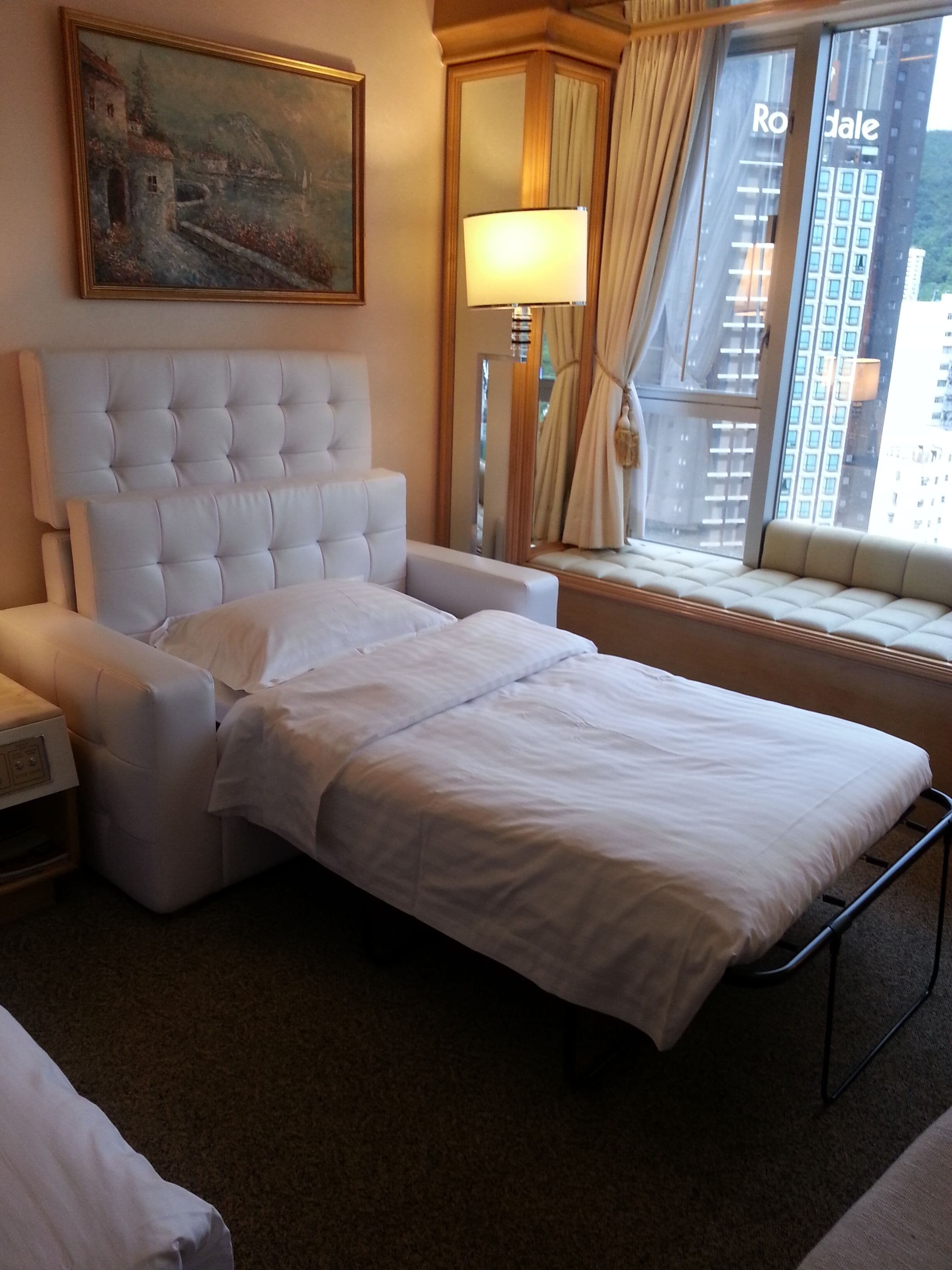 Quarto de hotel com uma cama de solteiro, mesa de cabeceira, abajur, janela com cortinas e um quadro na parede.