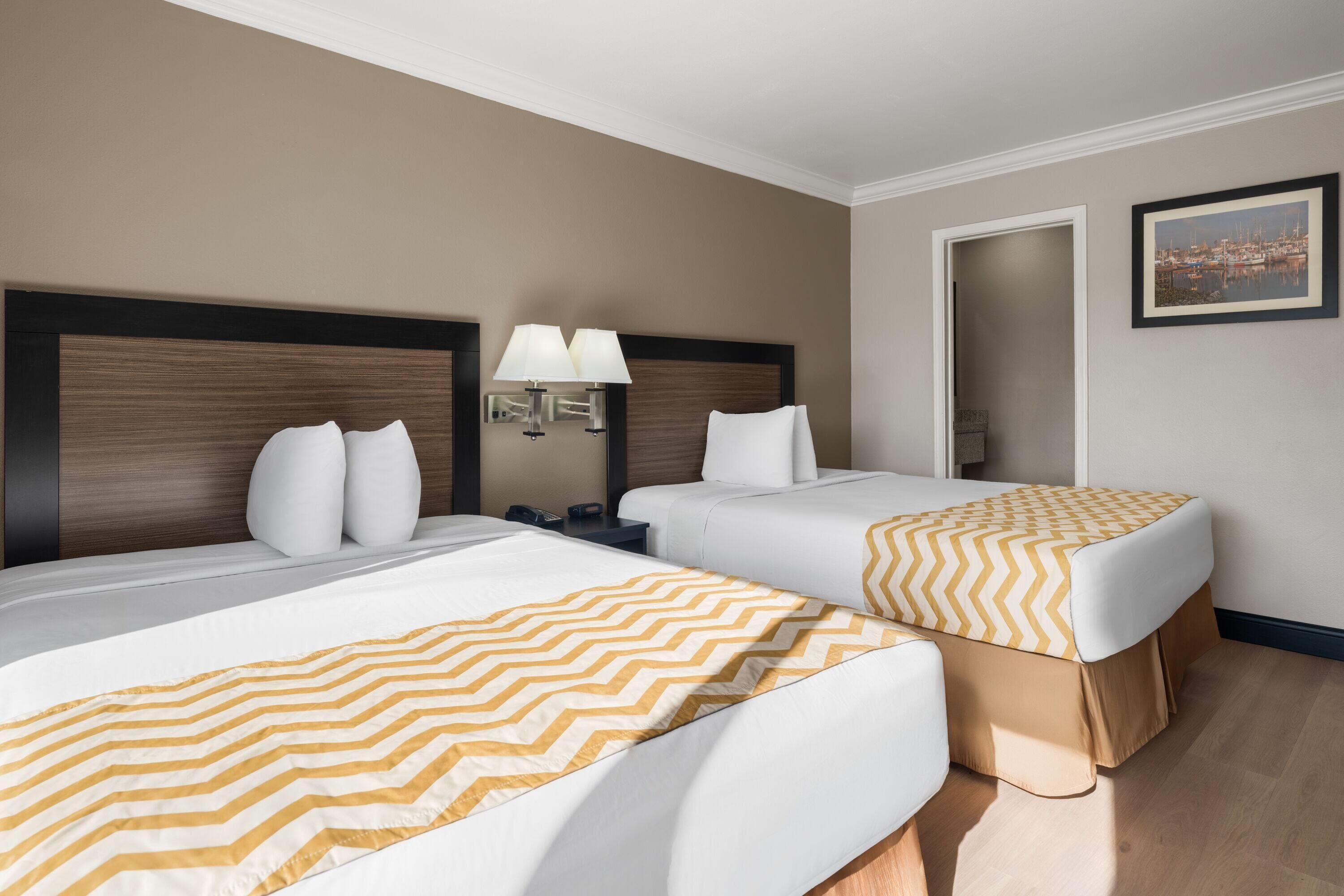 Quarto de hotel com duas camas, uma mesa de cabeceira, um abajur, um quadro na parede e uma porta que dá para outro cômodo.
