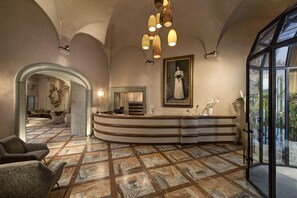 Uma área de recepção com piso de mármore, balcão de atendimento curvo, um grande retrato emoldurado e uma porta de vidro que dá para uma varanda com plantas.