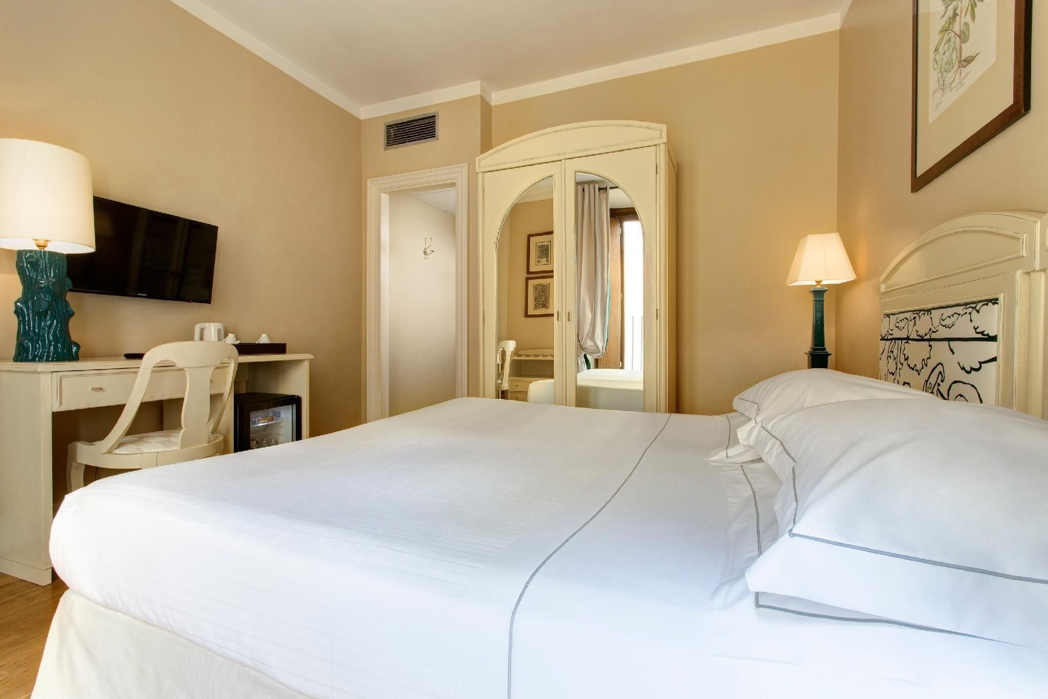 Quarto de hotel com cama, escrivaninha com cadeira, televisão, espelho e abajur.