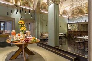 Uma área de refeições com uma mesa central exibindo frutas e bebidas, rodeada por assentos e um bar.
