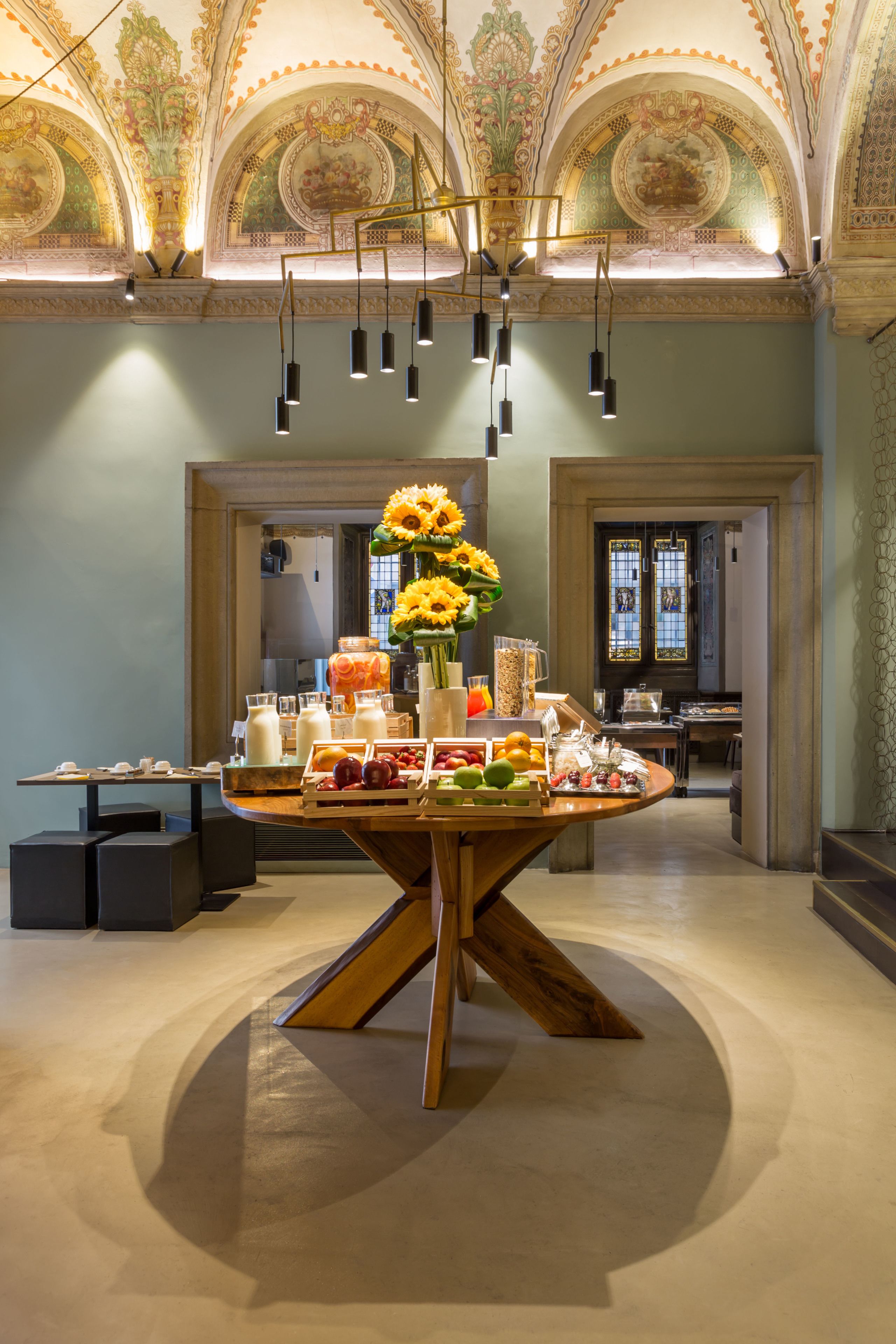 Um buffet de café da manhã com diversas frutas e bebidas, uma mesa de madeira com um arranjo floral central e um teto com desenhos elaborados.