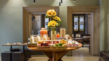 Um buffet de café da manhã com diversas frutas e bebidas, uma mesa de madeira com um arranjo floral central e um teto com desenhos elaborados.