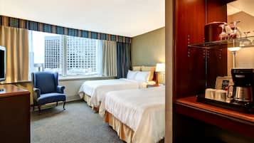 Quarto de hotel com duas camas, uma escrivaninha, uma cadeira, uma cafeteira e janela com vista para a cidade.